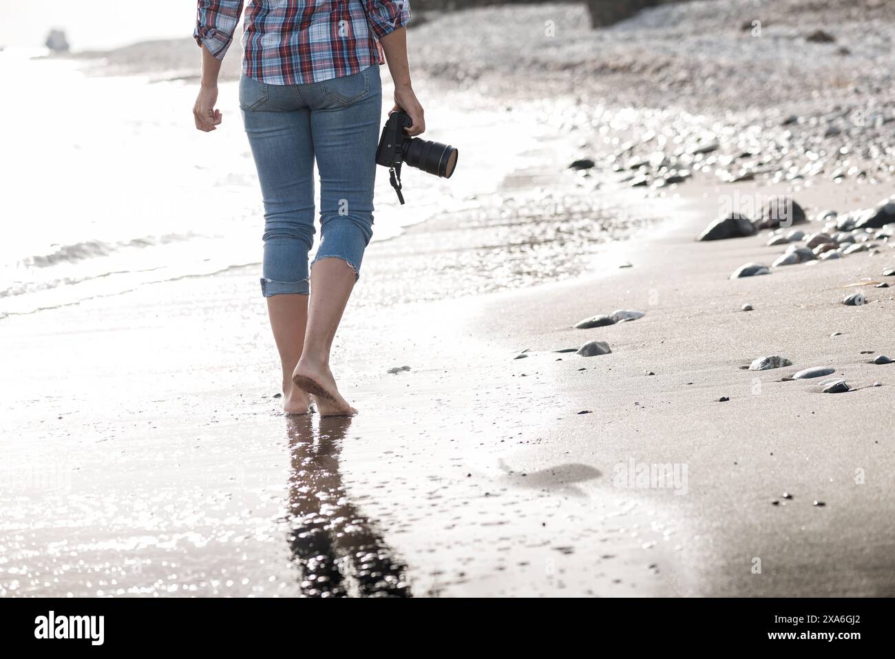 Woman holding a camera walks along a sunlit beach, leaving footprints ...