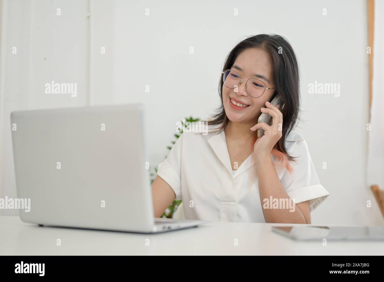 A young positive Asian woman is working remotely from a cafe, talking on the phone with her ...