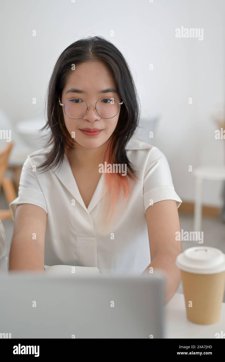 A young positive Asian woman wearing eyeglasses is working remotely ...