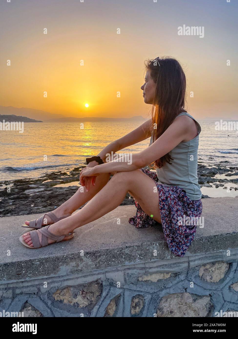 A woman sitting alone on stone ledge, watching ocean sunset in Greece ...