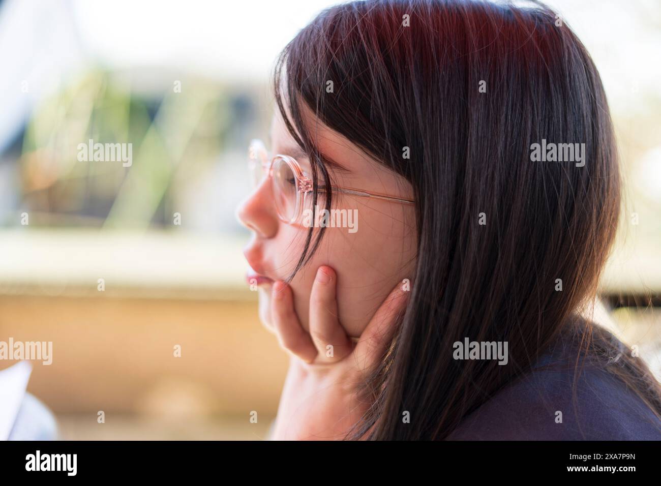 Sad dark-haired girl with glasses sitting at a table in the backyard ...
