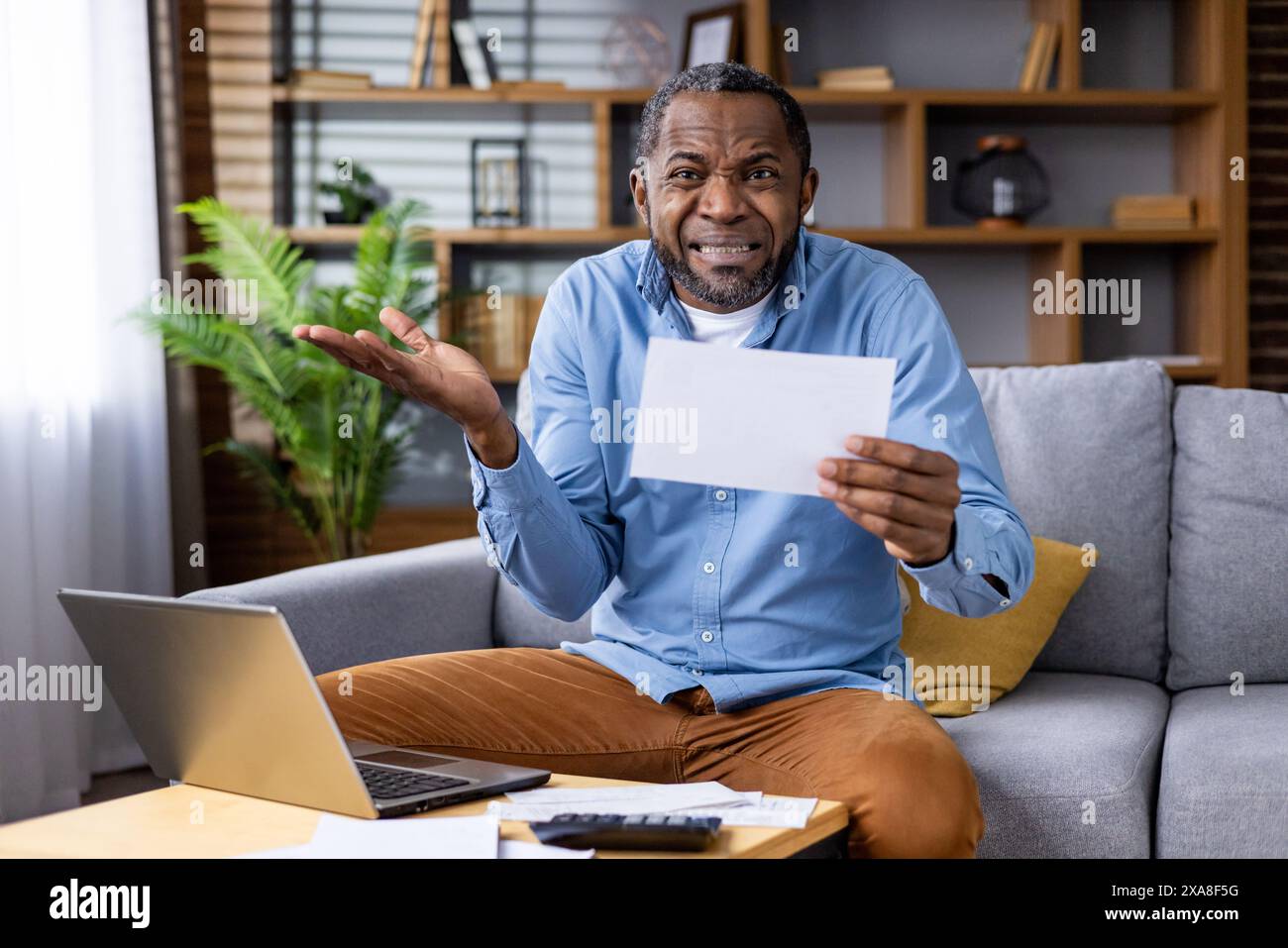 Frustrated man sitting on the couch looking at paperwork and gesturing ...