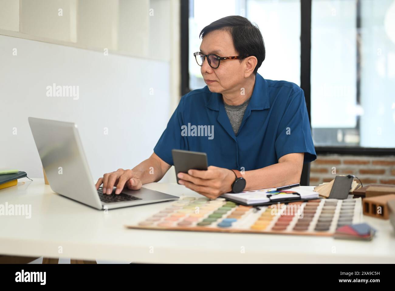 Mature interior designer using laptop and working with material and color samples at his ...