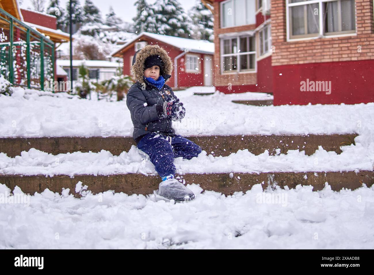 latino boy sitting in a warm hooded jacket playing in the snow at a ...