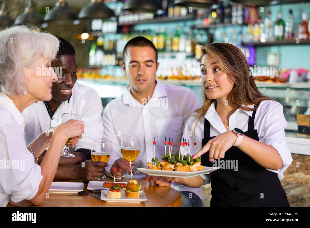 Cheerful Latin female waiter in apron serving pinchos for multiracial ...