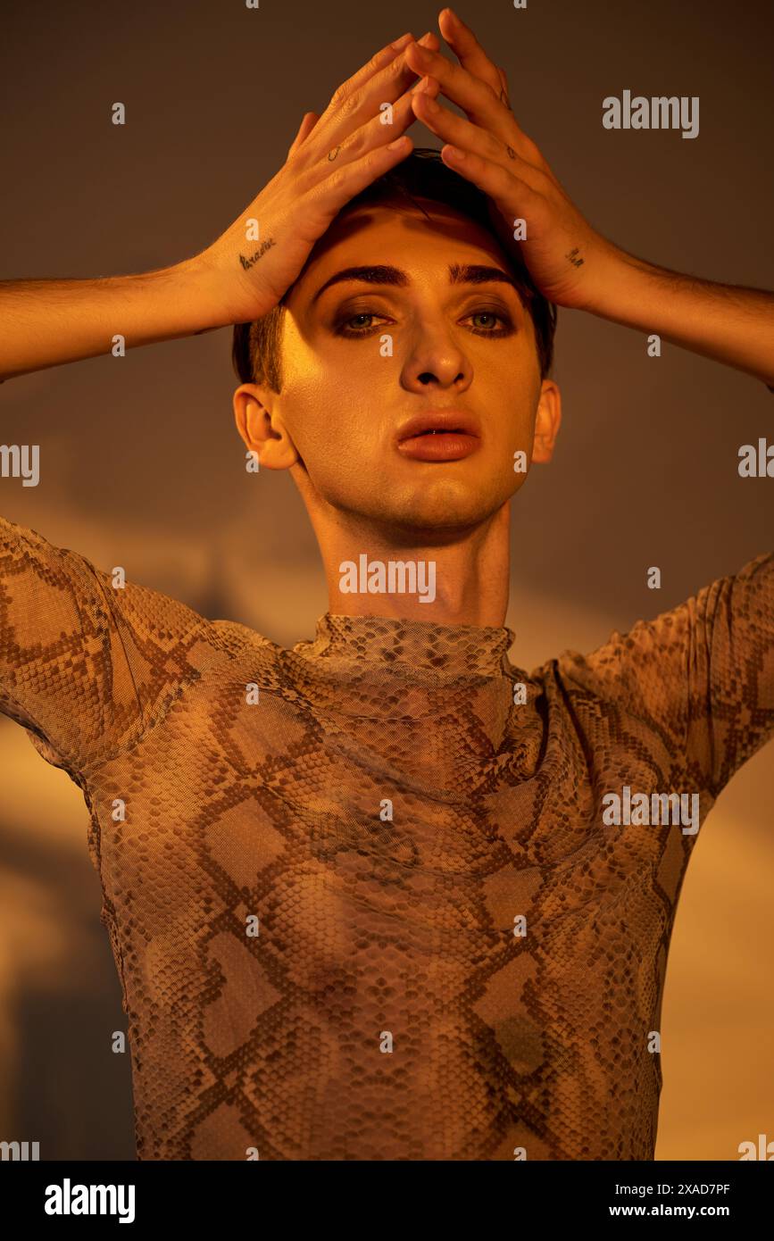 A young queer person in a snake skin top poses with hands on head in a ...
