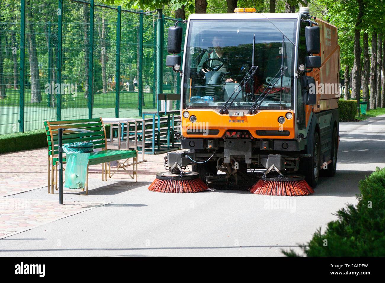 Street sweeper cleaning a park path near green benches and trash bins ...