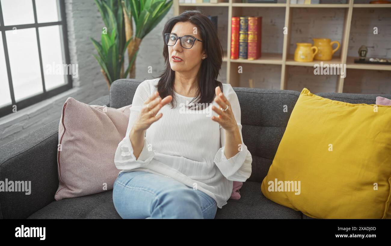 Portrait of a mature hispanic woman talking with hand gestures, seated on a grey couch in a well ...