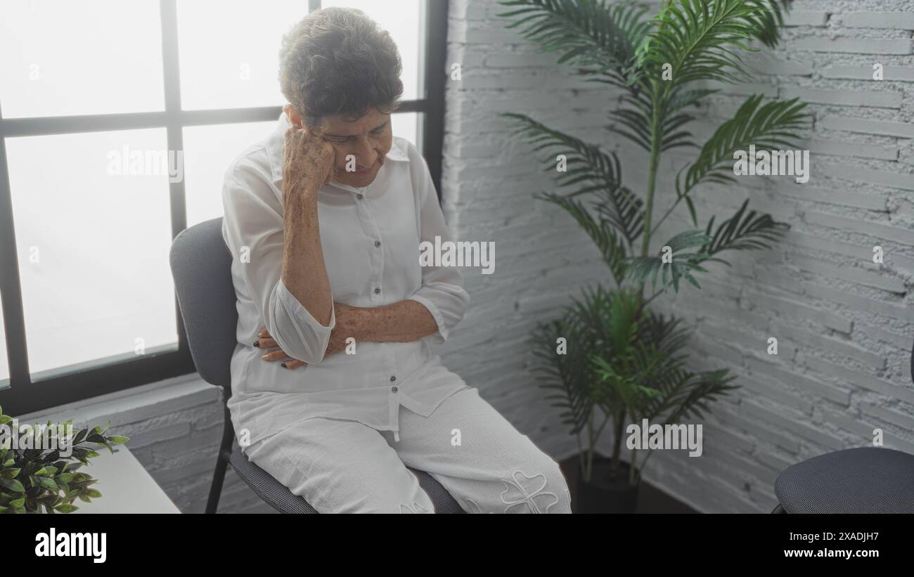 Elderly woman with short hair sits pensively in a white shirt and pants ...