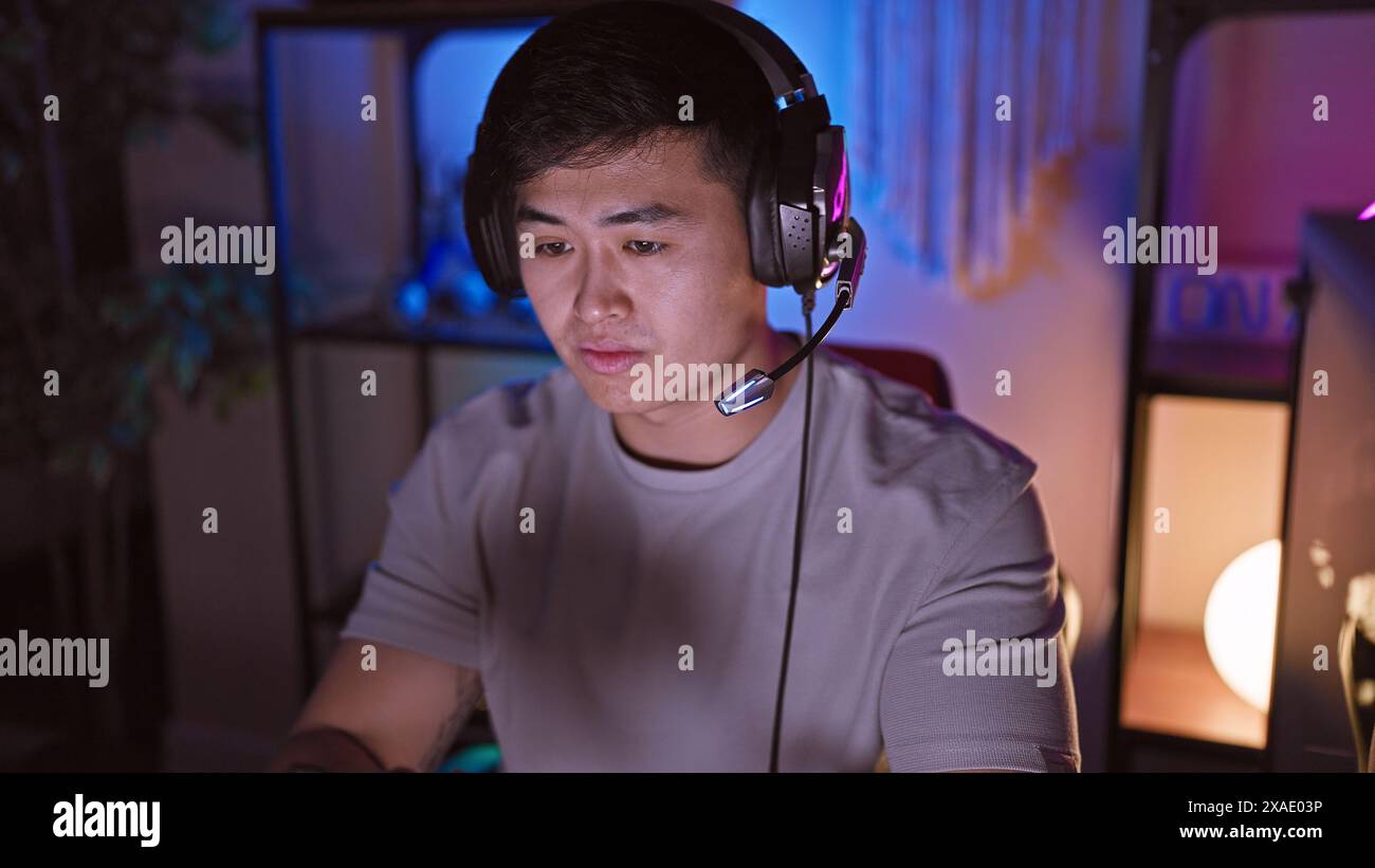 Asian man with headphones focused while gaming in a dark room lit by ...