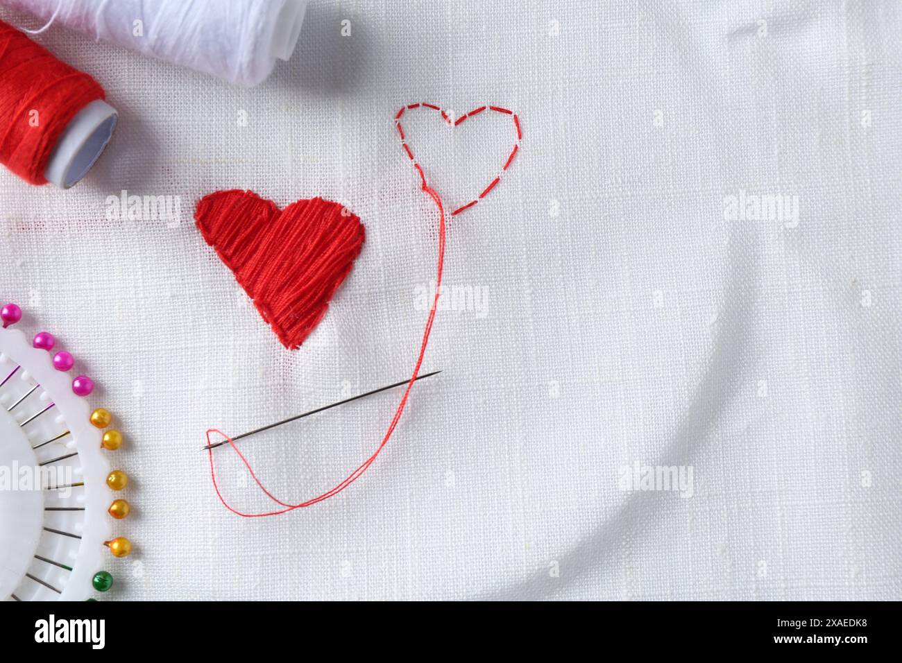 Embroidered red hearts and needle on white cloth, top view. Space for ...