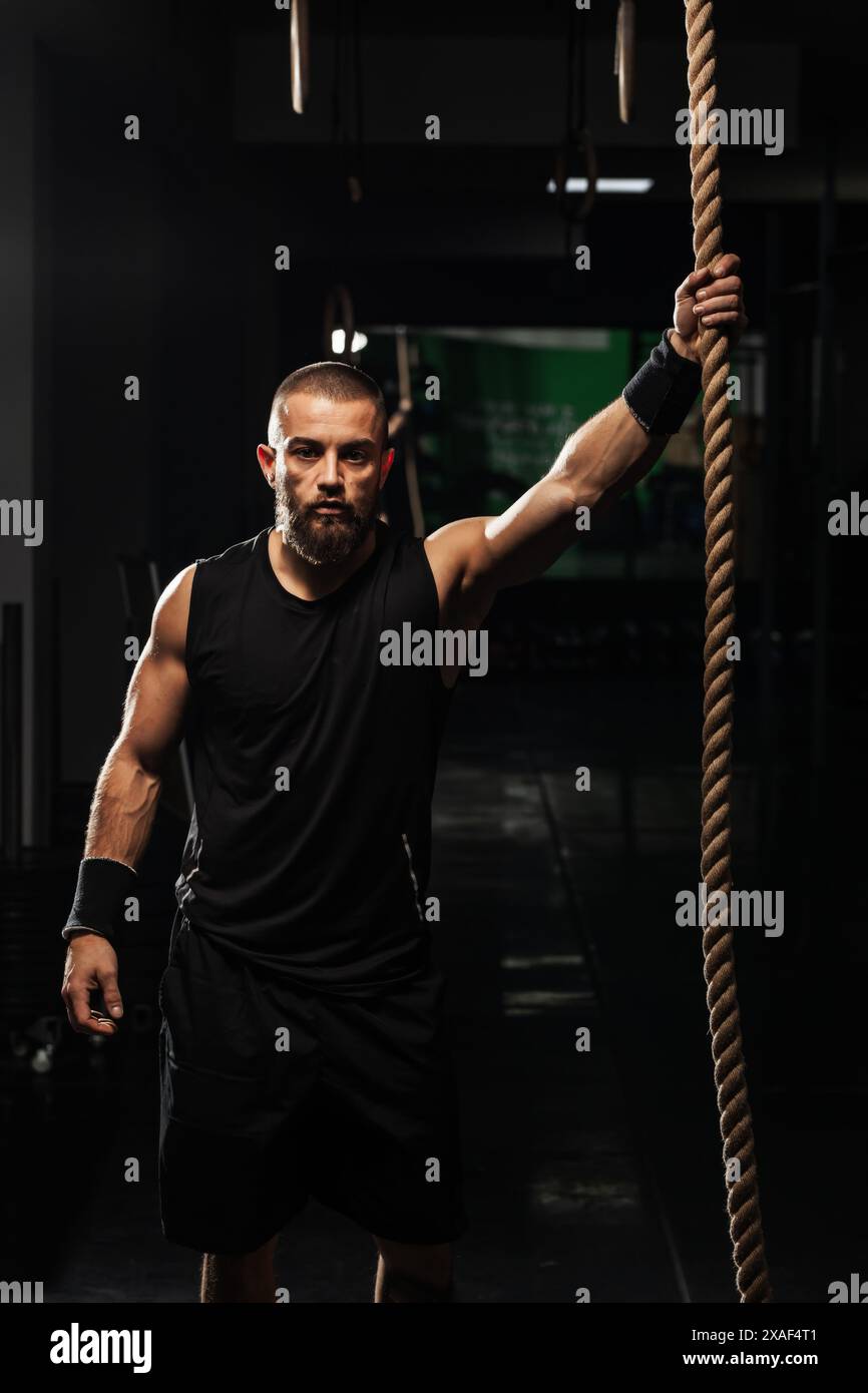 Strong male fitness enthusiast prepares to climb a rope in a dimly lit ...