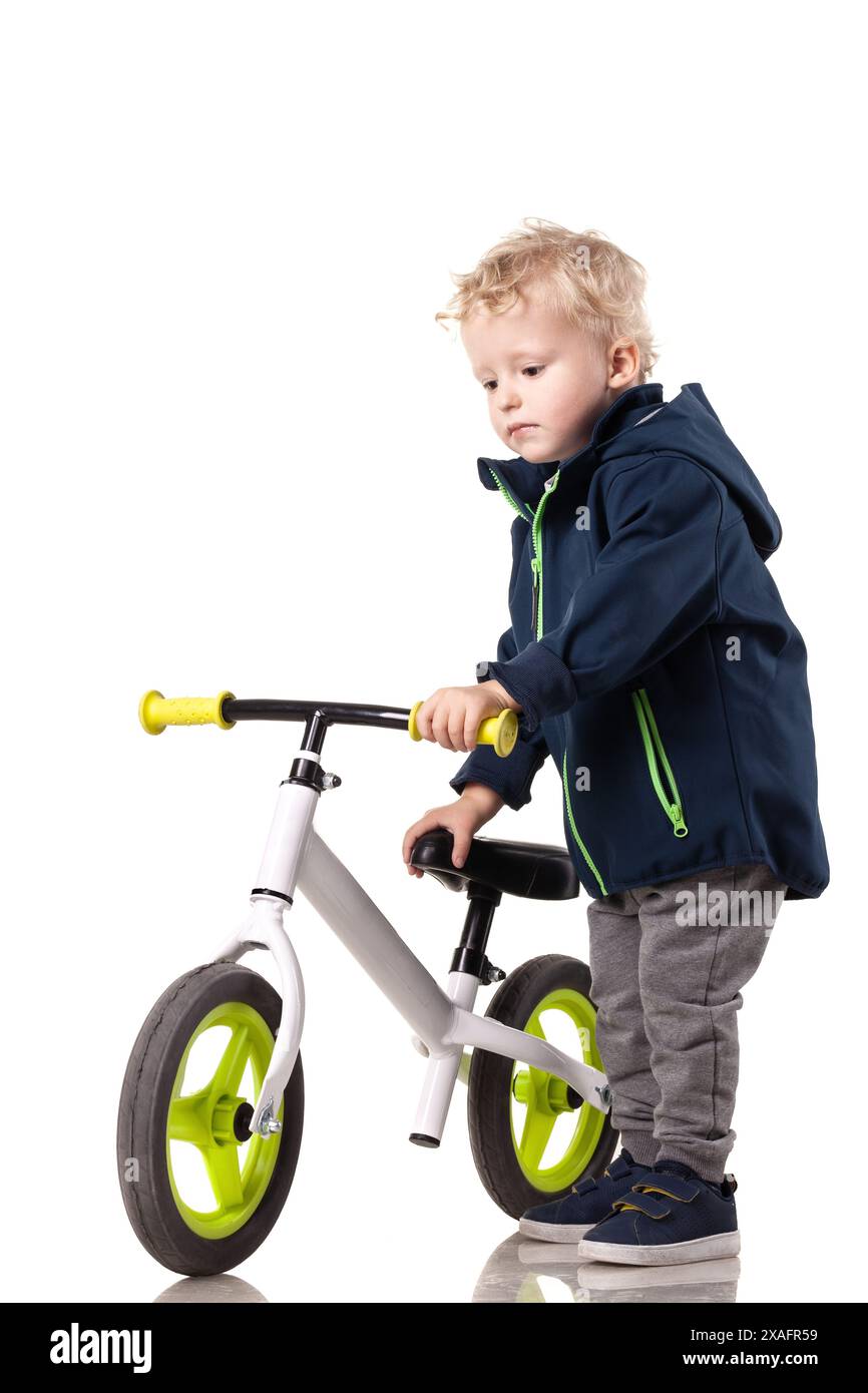 Blonde child in a blue jacket stands ready to ride a white balance bike ...