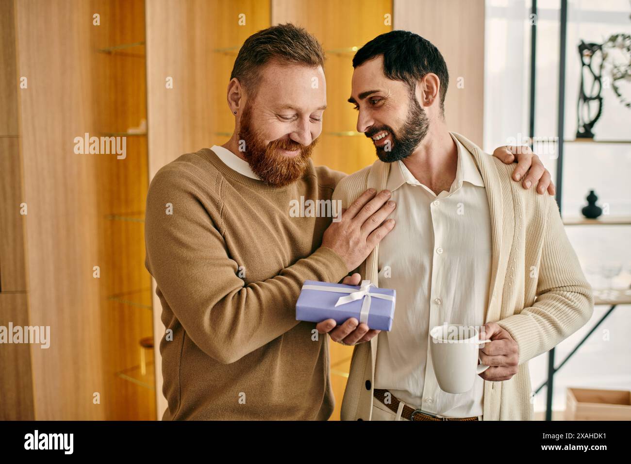 Two men joyfully exchange gift boxes in a modern apartment, celebrating ...