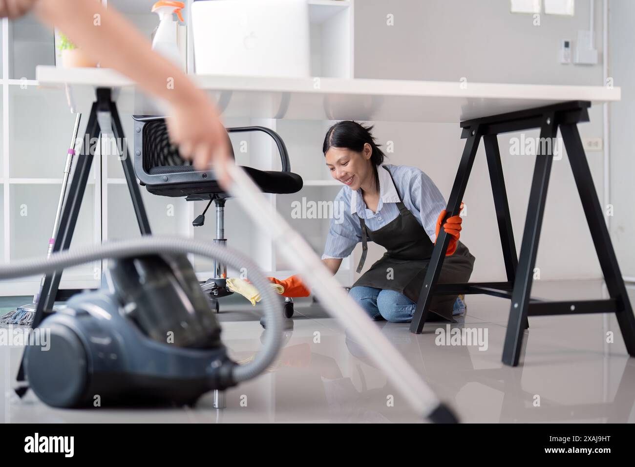 Female office cleaner wiping floor under desk with vacuum in foreground ...