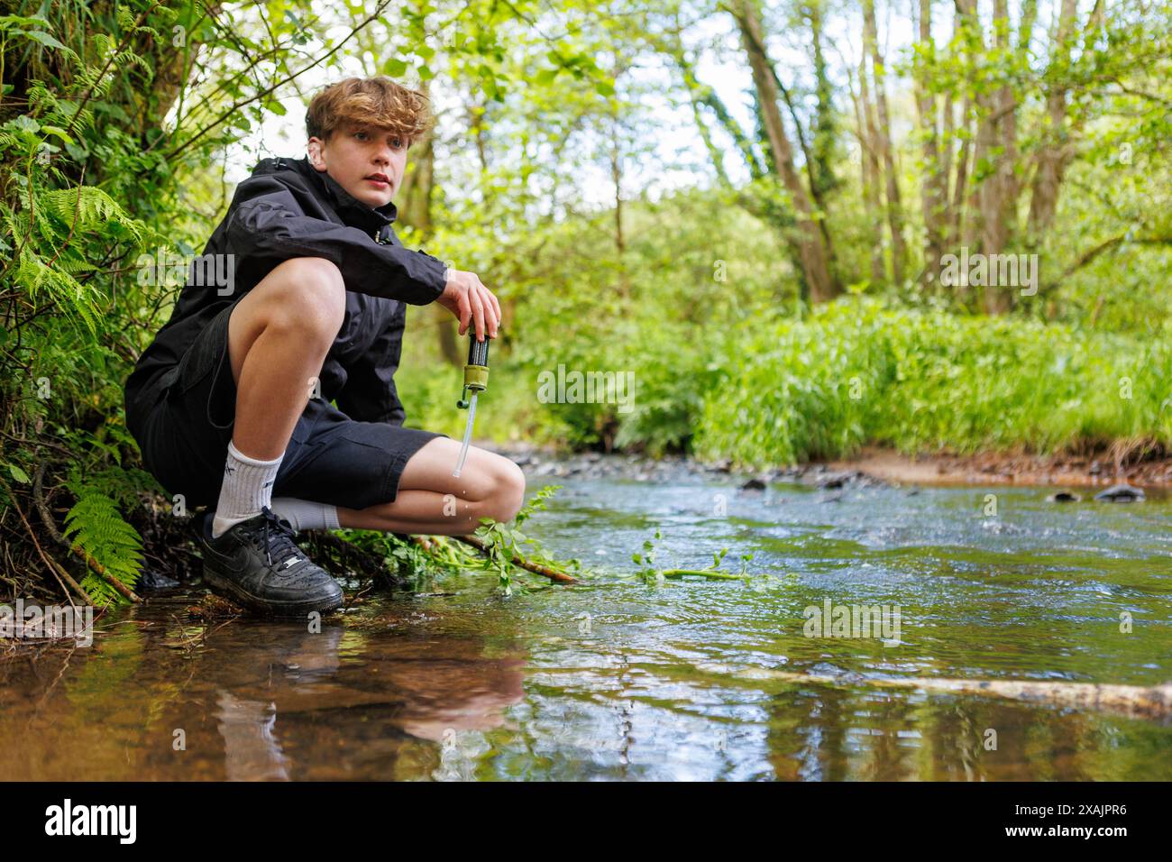 A young teenage boy crouches down next to a small stream to use his ...
