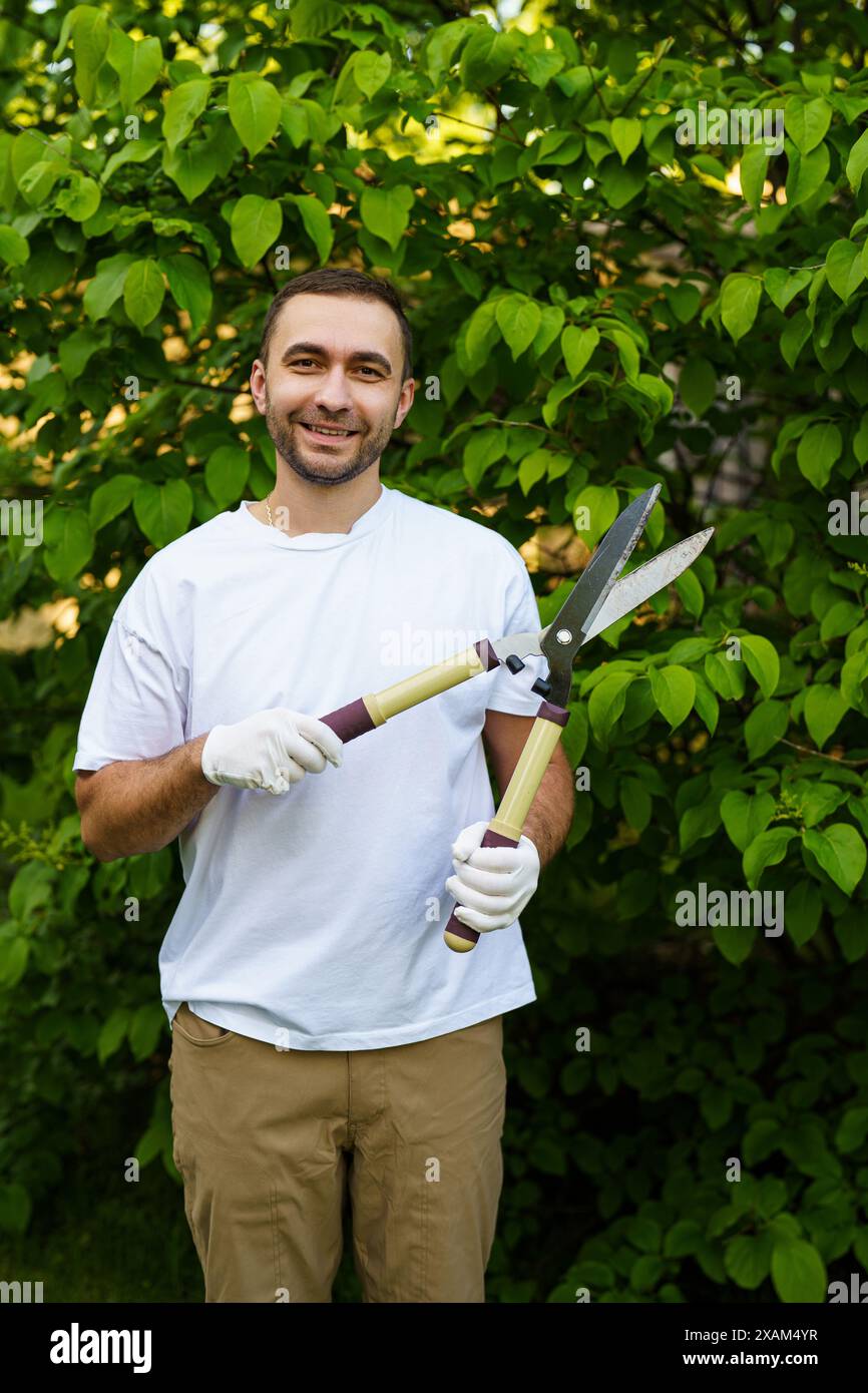Man gardener in gloves doing maintenance work, pruning trees. Green fence during sunny summer ...