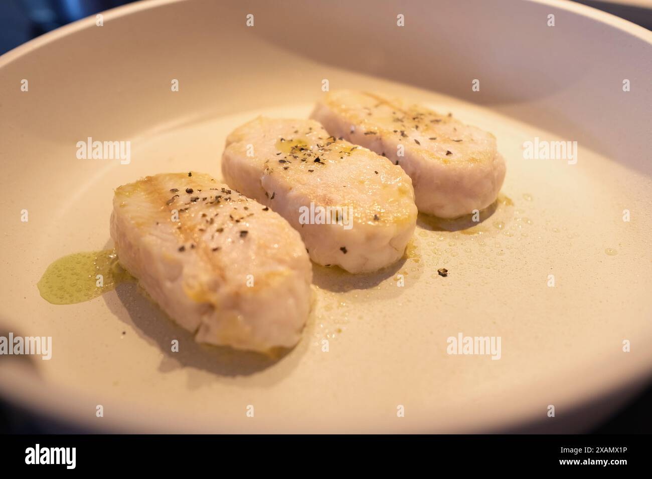 Three fresh hake loins are cooked in a white granite pan with extra ...