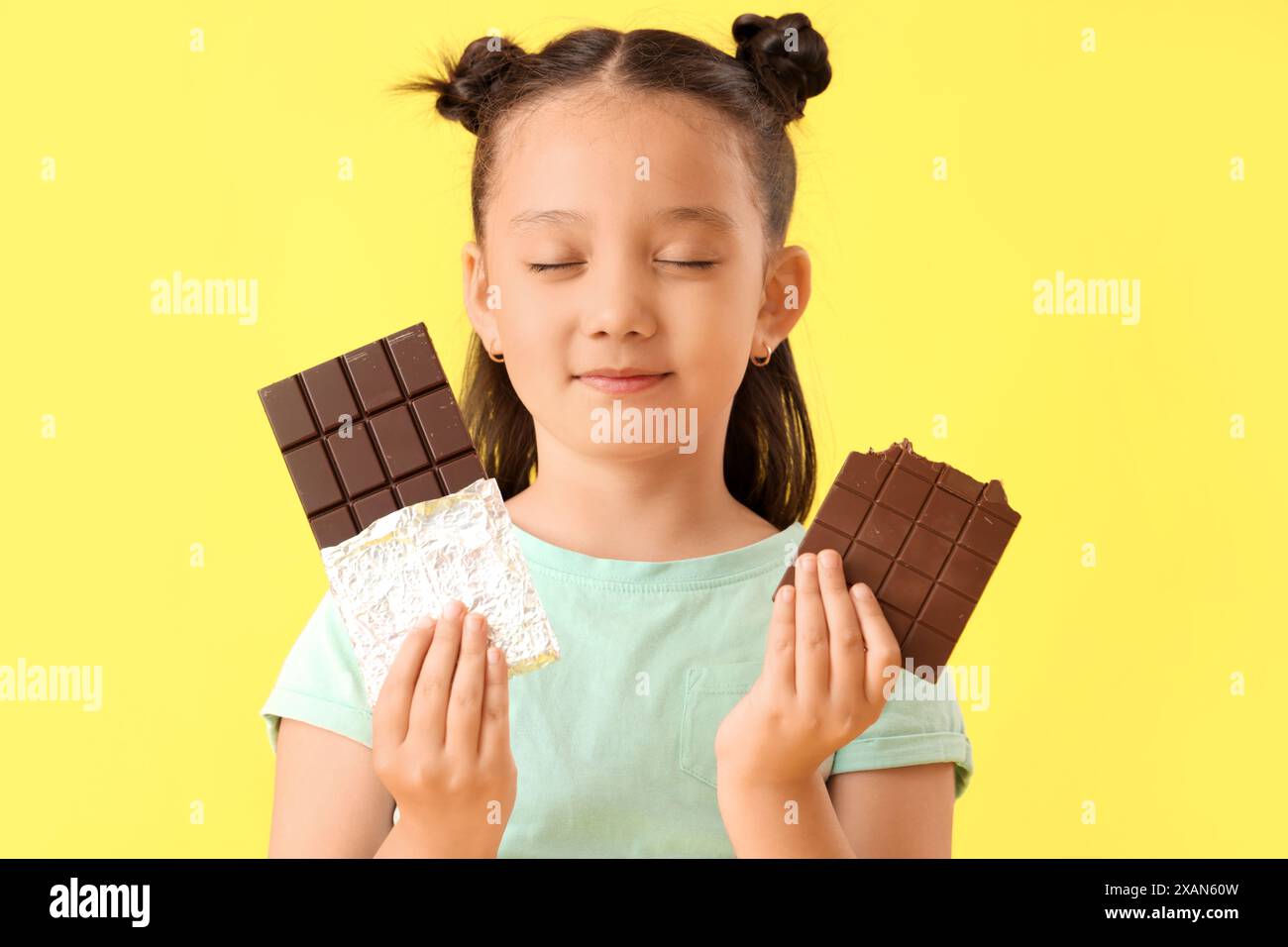 Cute little Asian girl with chocolate bars on yellow background ...