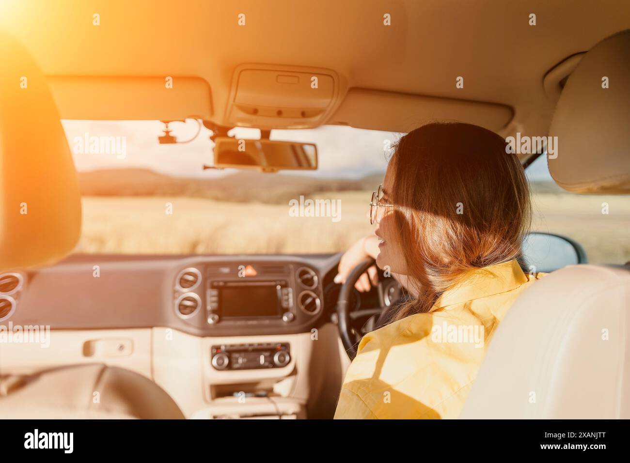 A woman in a yellow shirt is driving a car. She is wearing glasses and ...