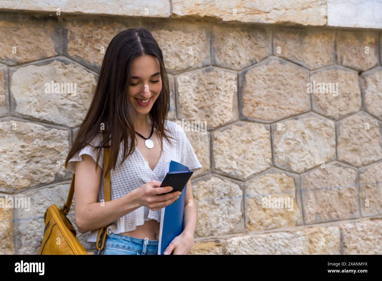 Relaxed young college student leaning against a wall, checking messages ...