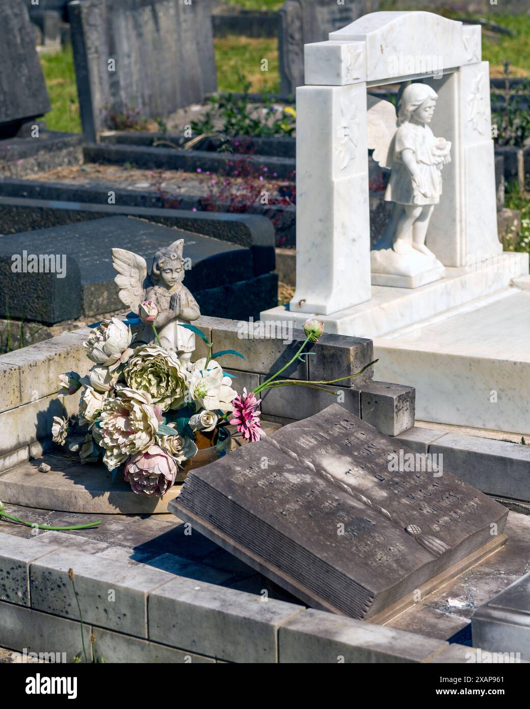 A cherub prays over a floral tribute on a grave marker. With a second ...