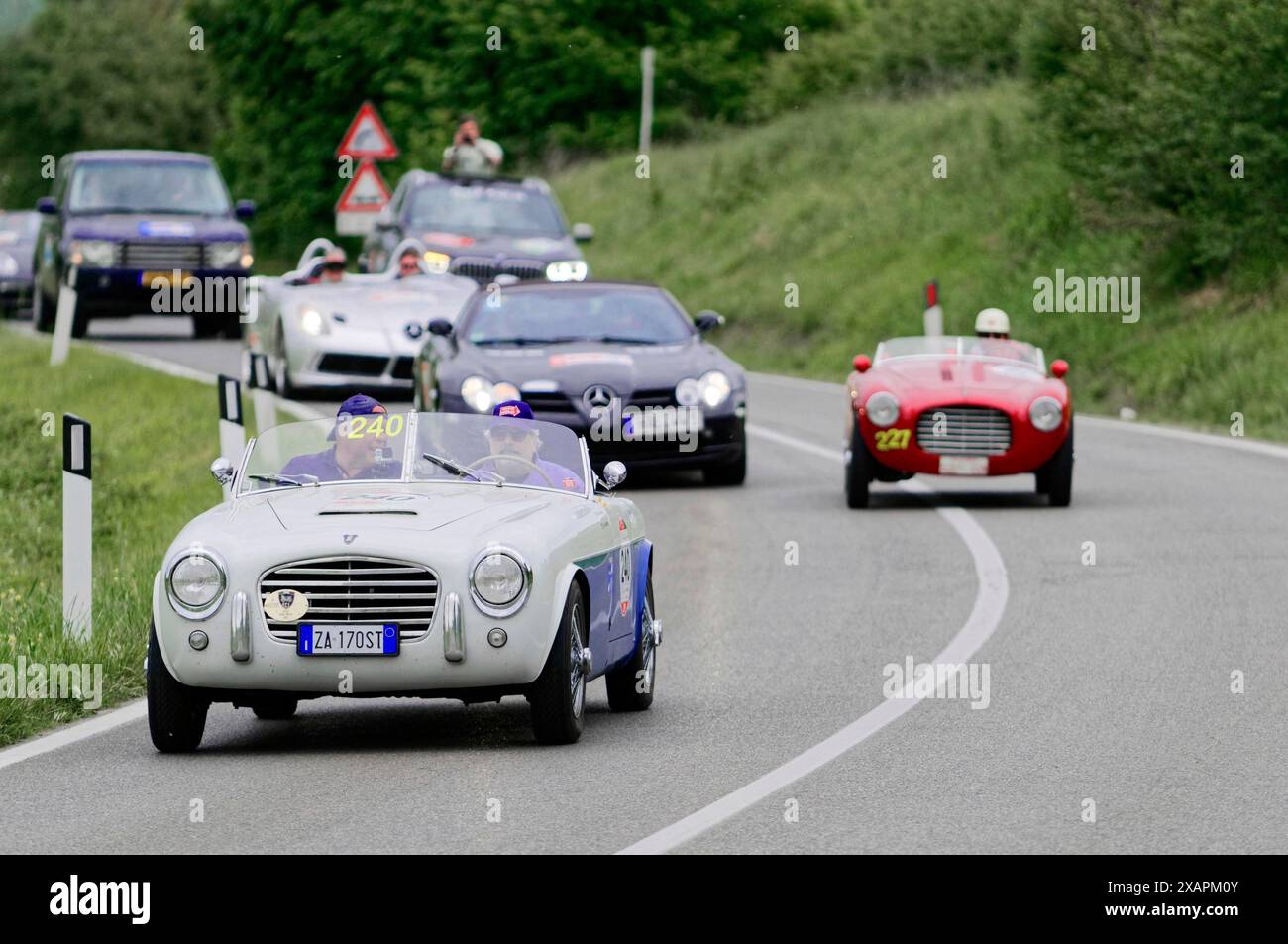 A convoy of vintage racing cars drives on a winding country road ...