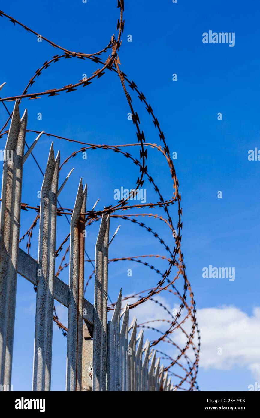 Rusty barbed wire on top of spiky metal fence. The blue sky is visible ...