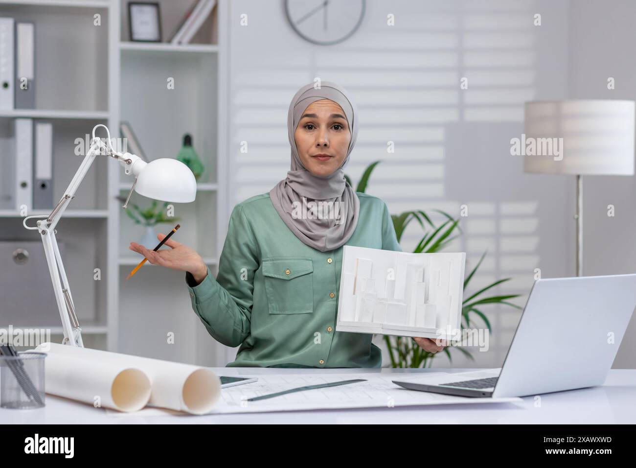 A confused female architect in a modern office holding a city model ...