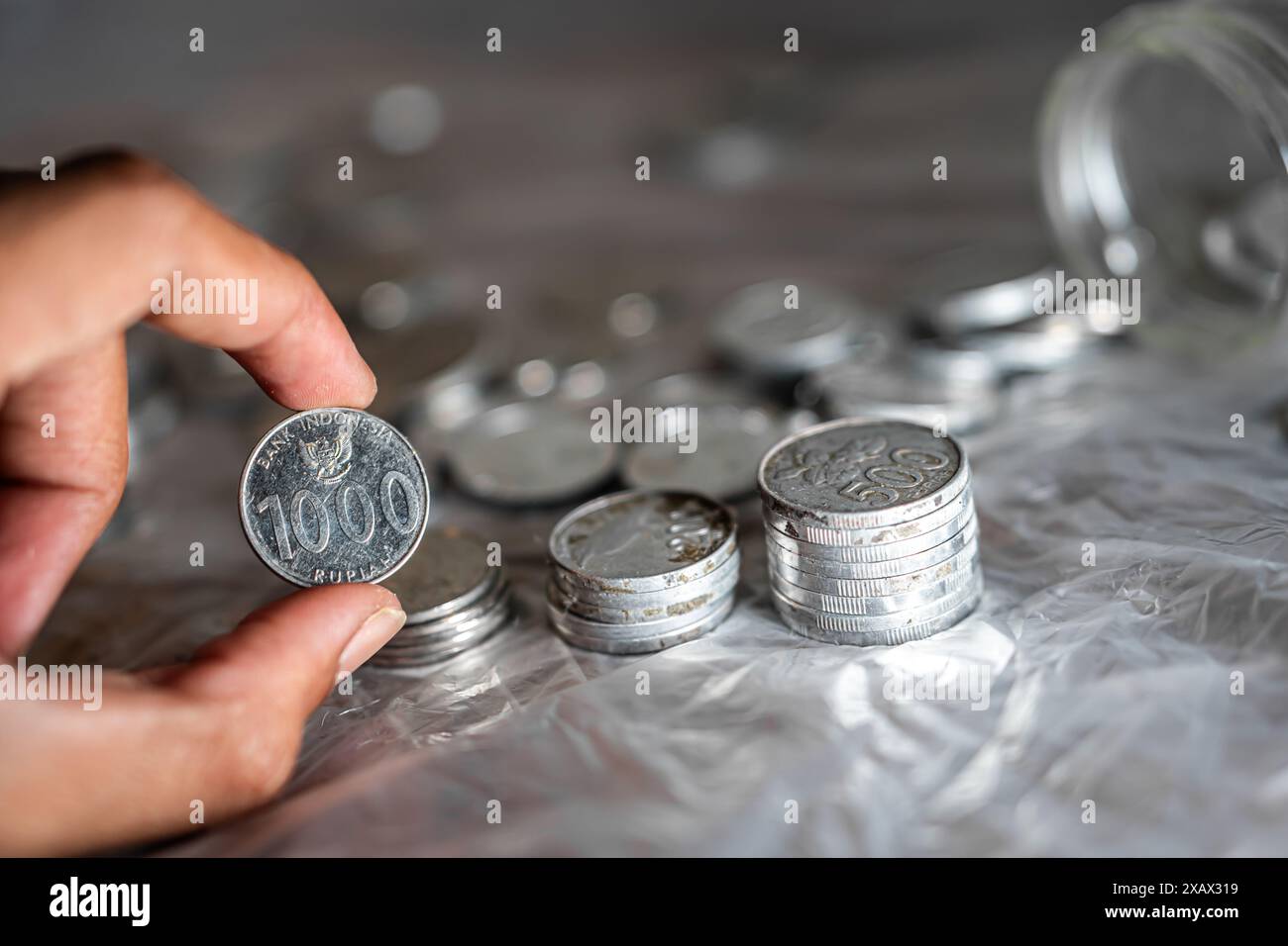 A close-up of a hand holding a 1000 Rupiah coin with stacks of similar ...