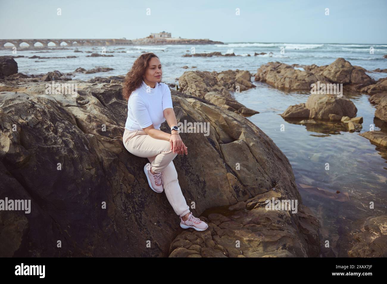 Happy woman sits on rugged coastal rocks, looking thoughtful and ...