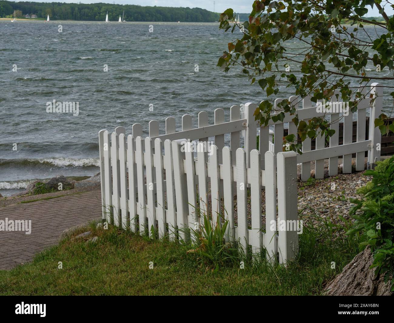 A white fence borders a garden next to a lake, surrounded by green ...