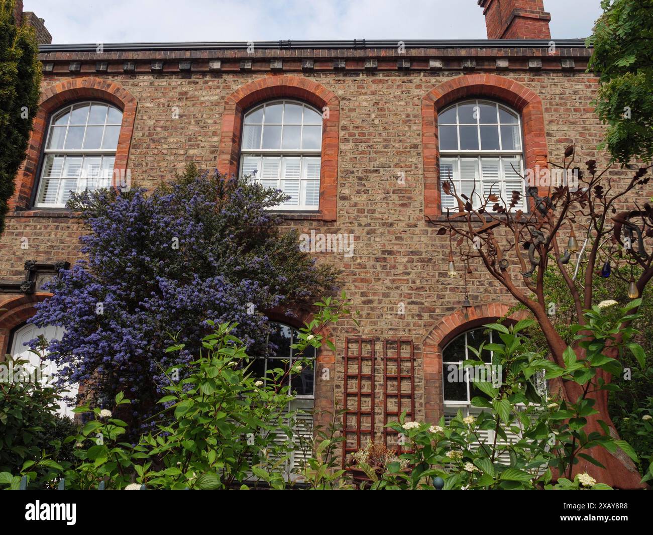 A historic brick building with large windows surrounded by lush gardens ...