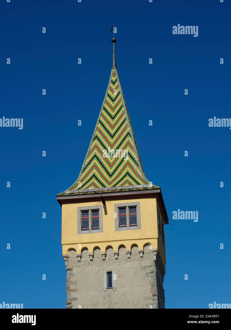 A medieval tower with a colourful tiled roof, in front of a clear sky ...