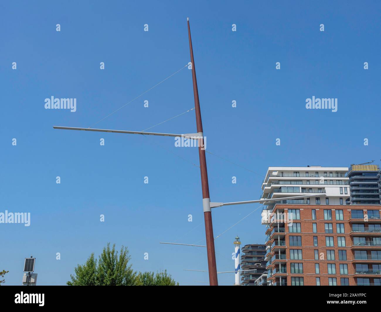 A tall sailing mast in front of modern buildings under a clear blue sky ...