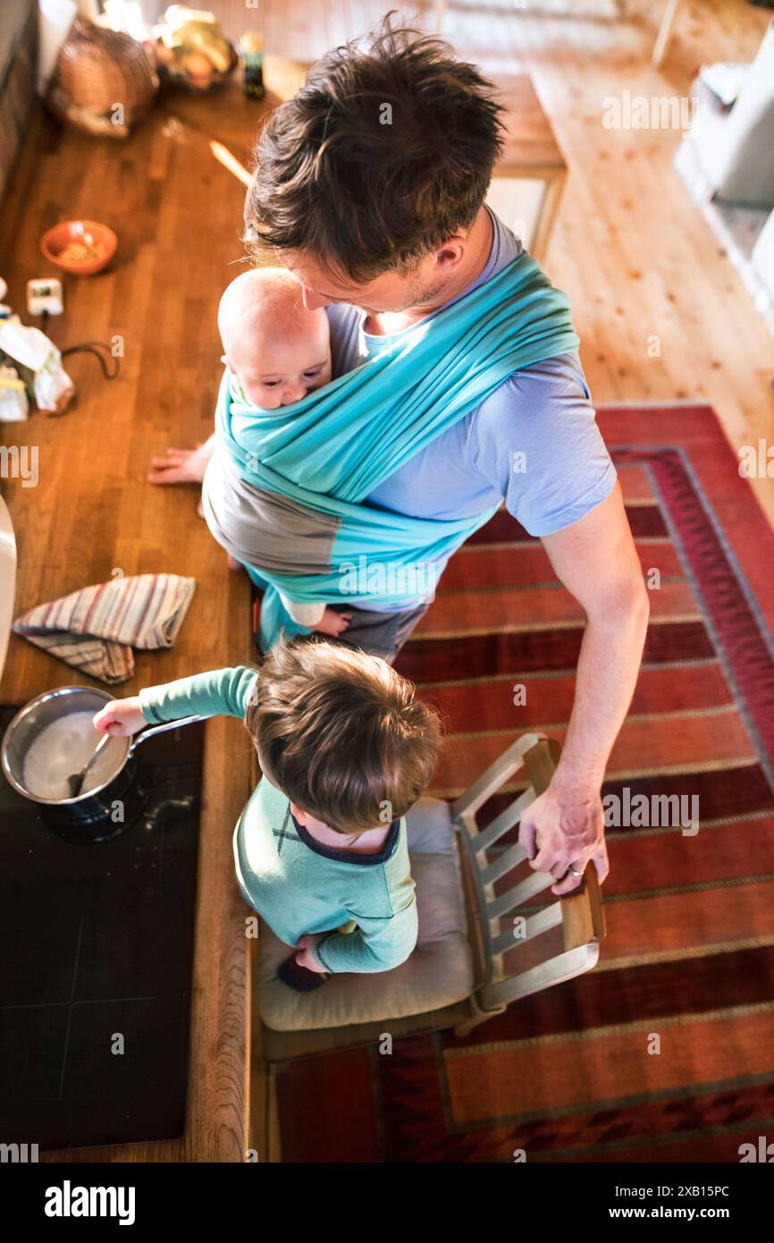 Father holding small baby in baby sling. Older brother stirring ...
