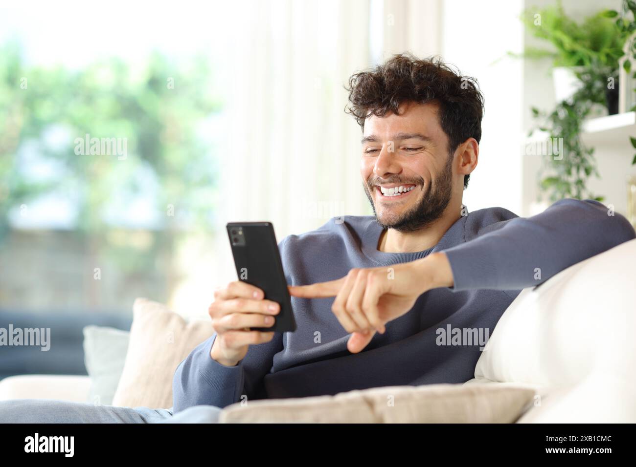 Happy man using smart phone sitting on a couch in a house interior ...