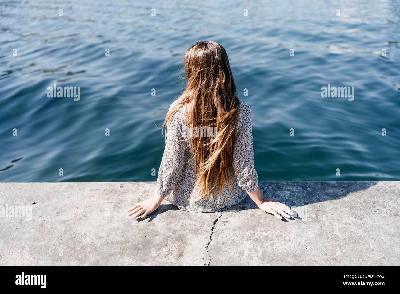 A woman is sitting on a ledge by a body of water, wearing a dress with ...