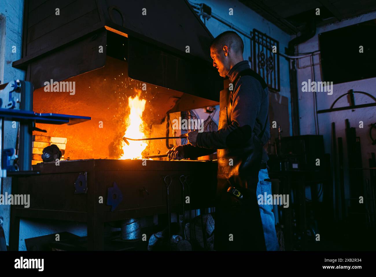 A male blacksmith forges metal with a hammer at an anvil amidst the fiery glow of a traditional ...
