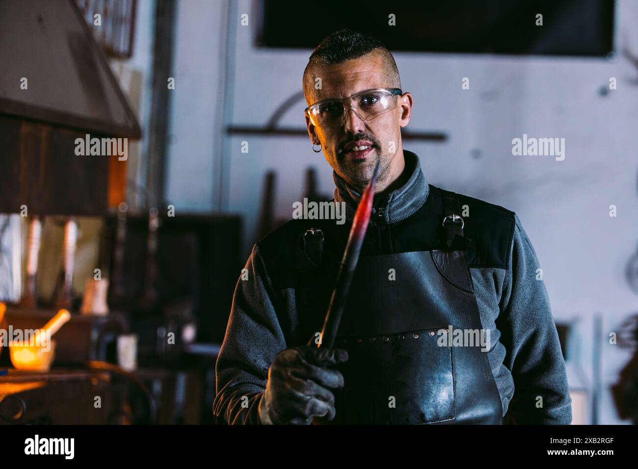 Skilled male blacksmith in a workshop holding a glowing hot forged iron rod, with protective ...