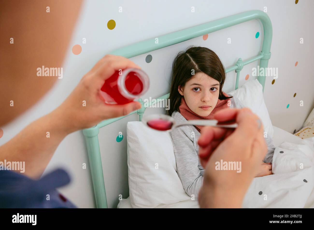 An adult hands over a spoonful of medicine to a young girl sitting up ...