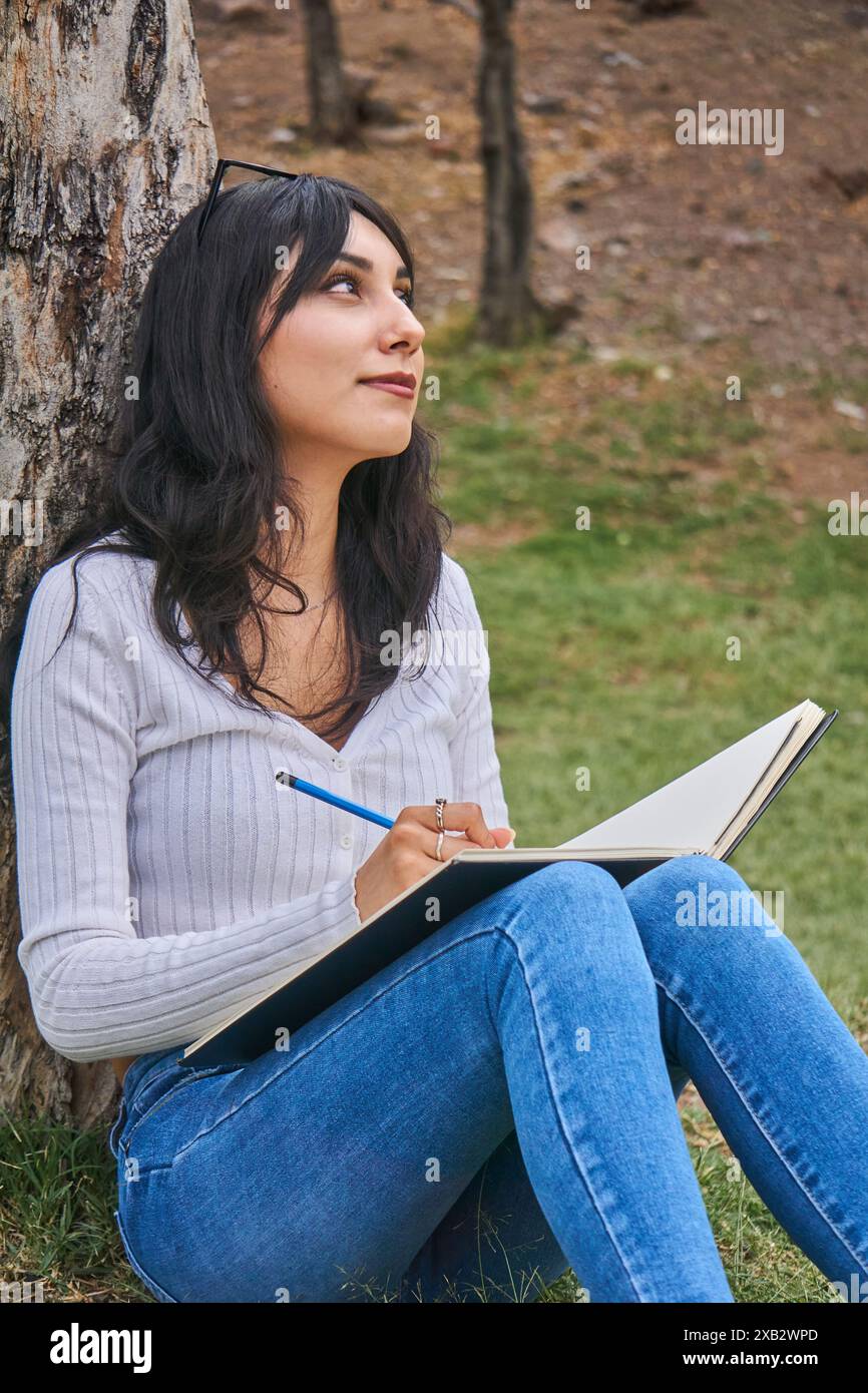 A contemplative woman sits by a tree in a tranquil park setting ...