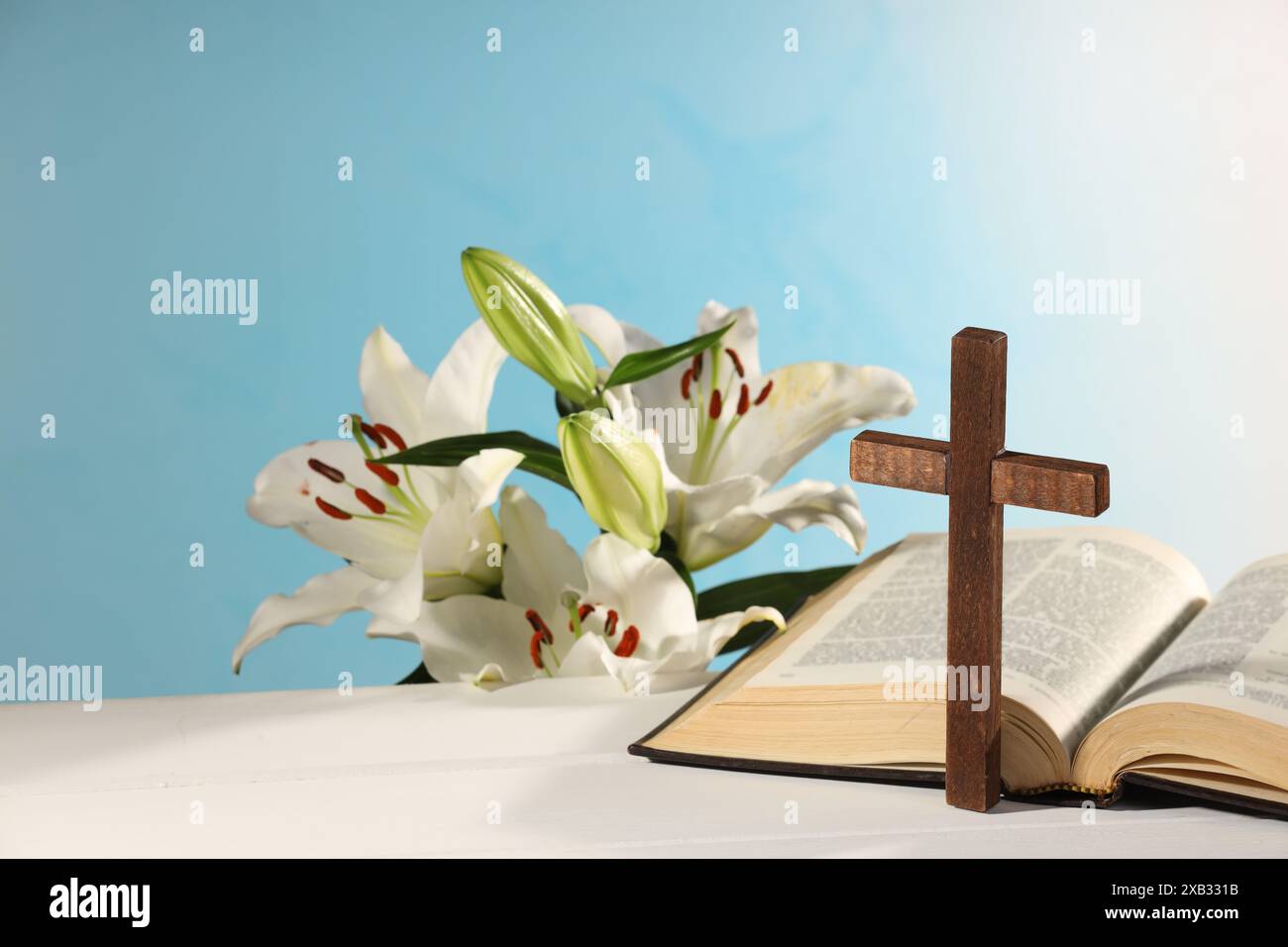 Wooden cross, Bible and beautiful lily flowers on white table against ...