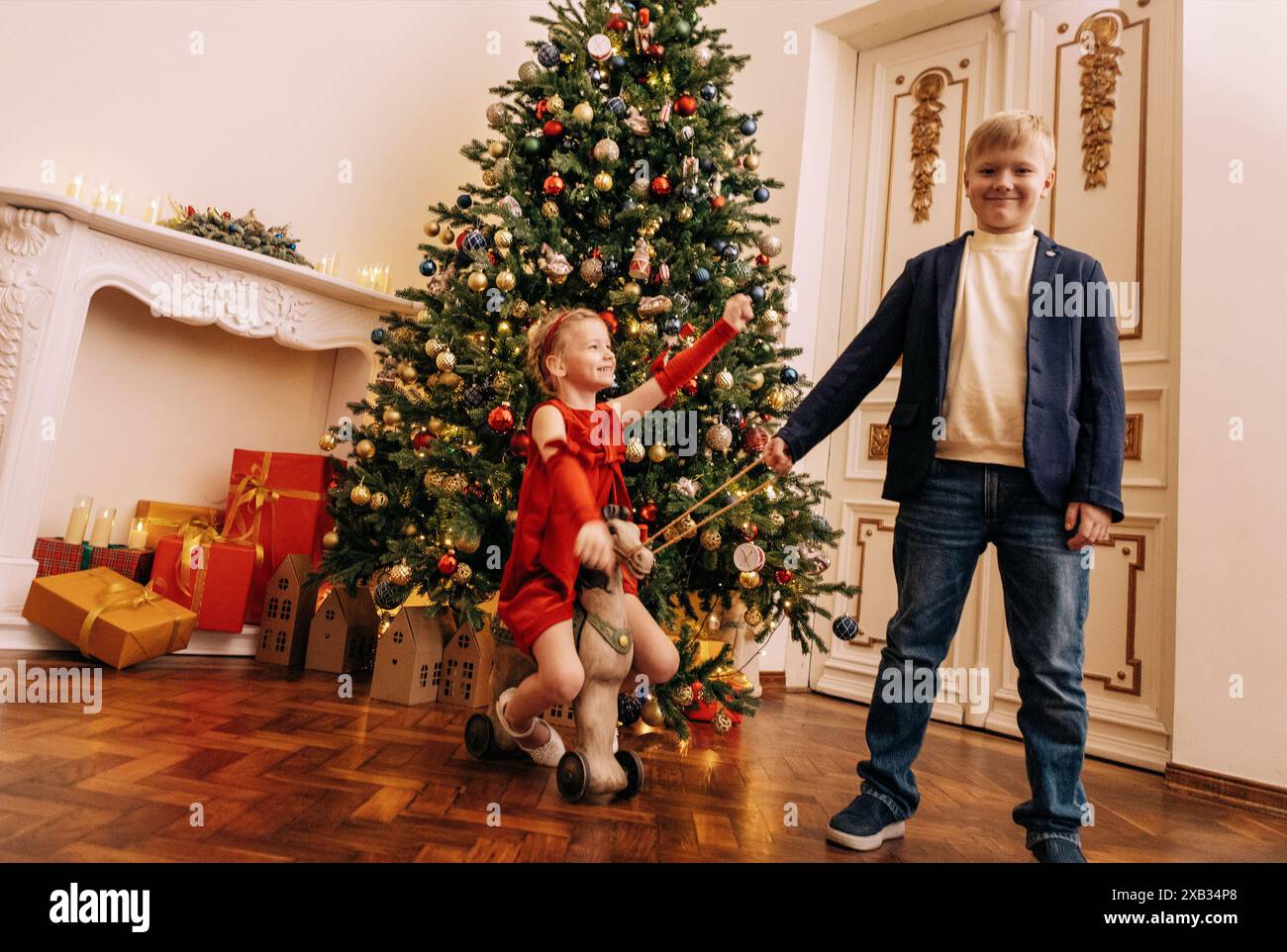 Smiling siblings in fancy clothes are playing near Christmas tree at ...