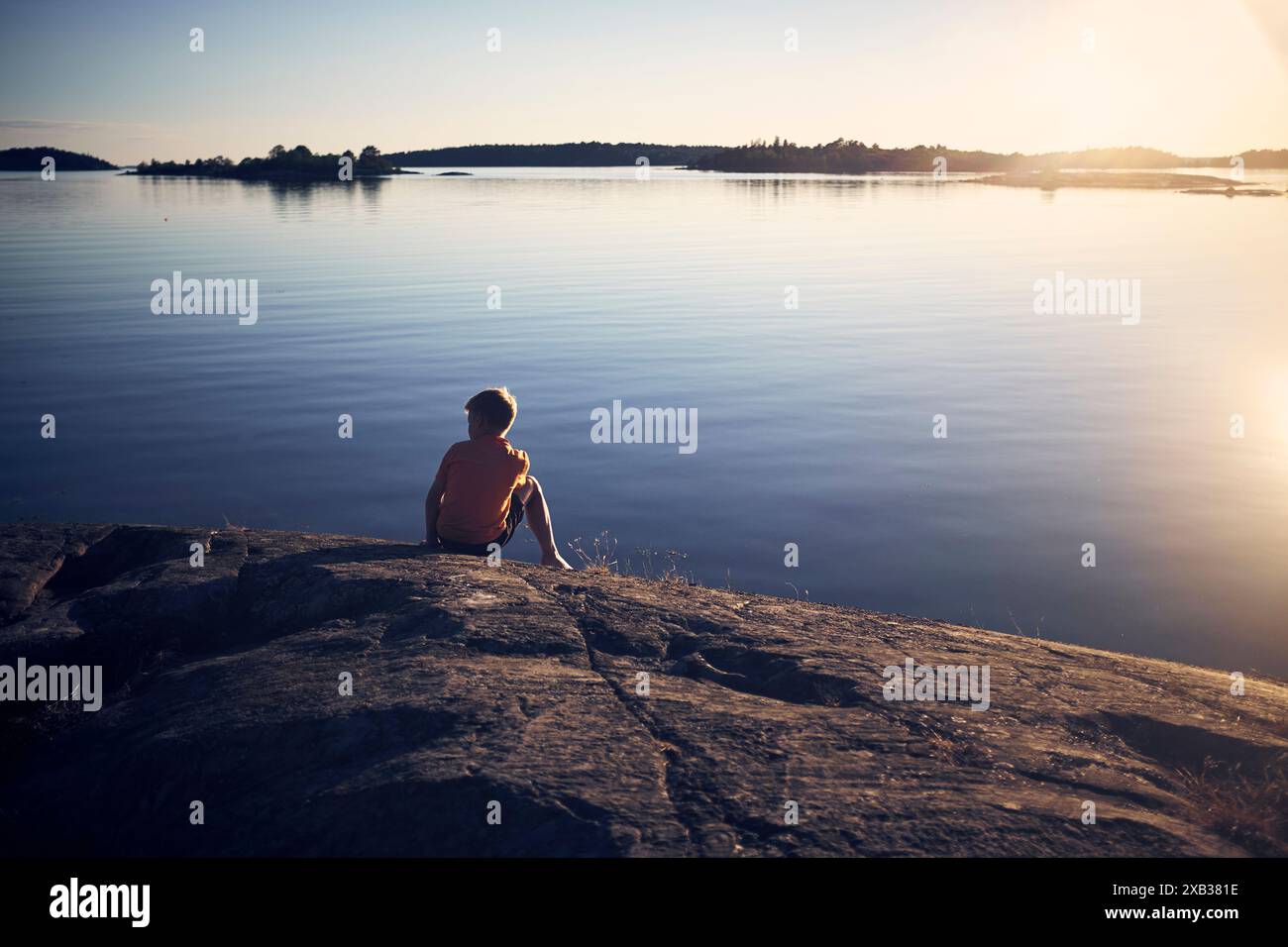 Rear view of boy sitting on rock's edge at lakeshore during sunset ...
