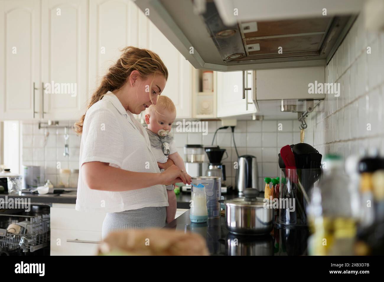 Woman carrying toddler son while preparing milk bottle at kitchen ...