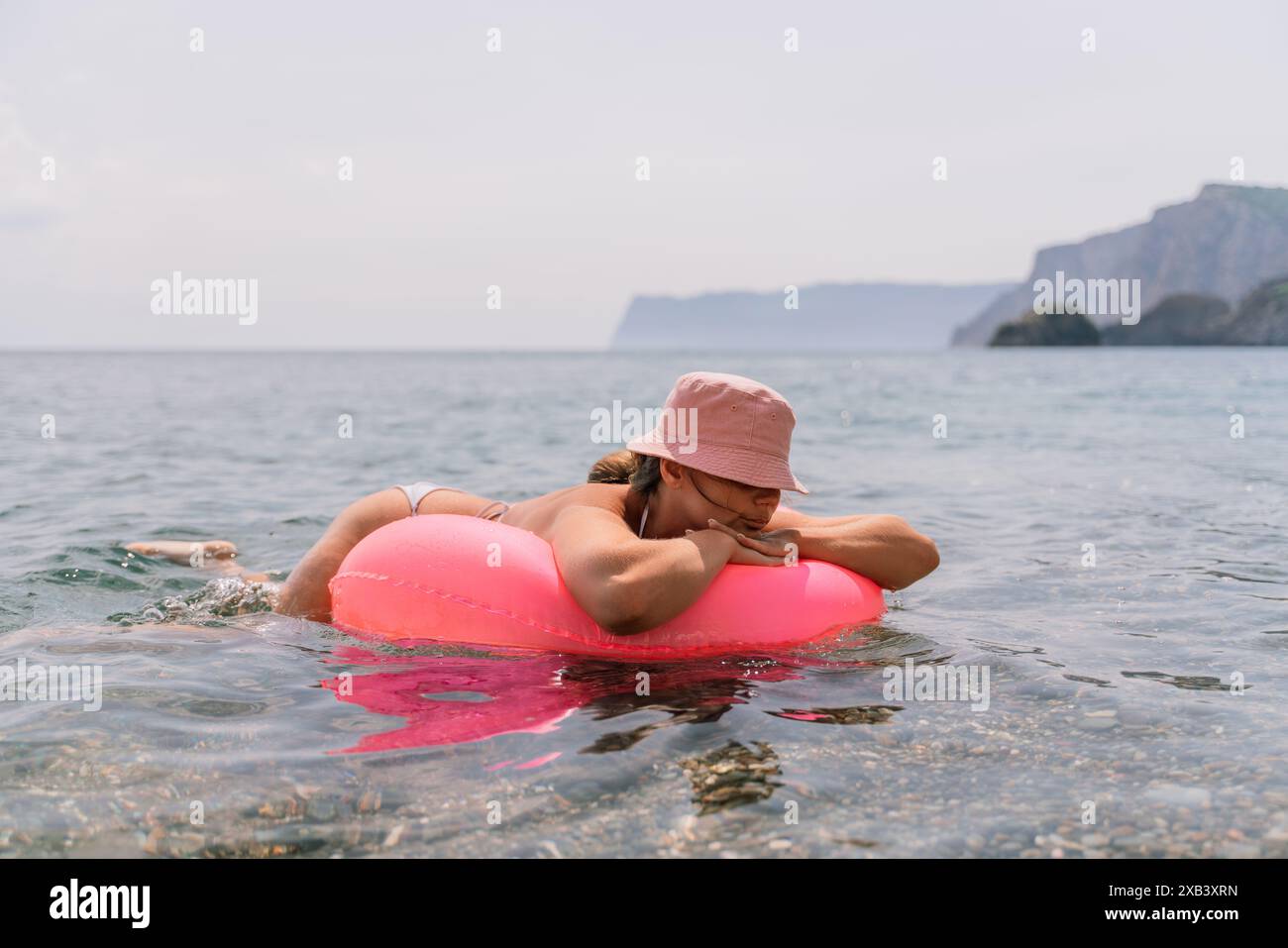 A woman is laying on a pink inflatable raft in the ocean. The scene is ...