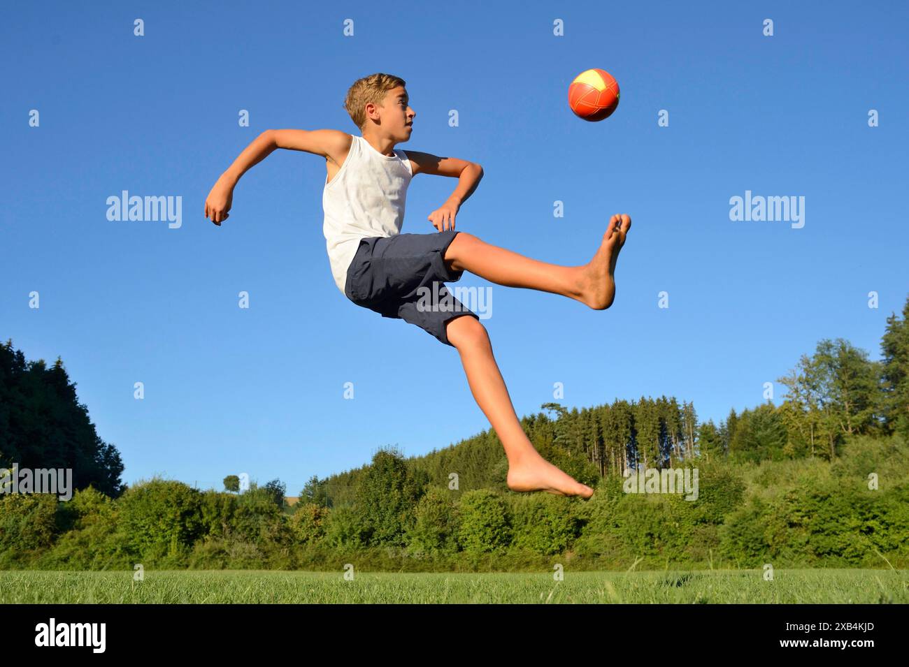 A boy mid-air kicking a soccer ball in a grassy field under a clear ...