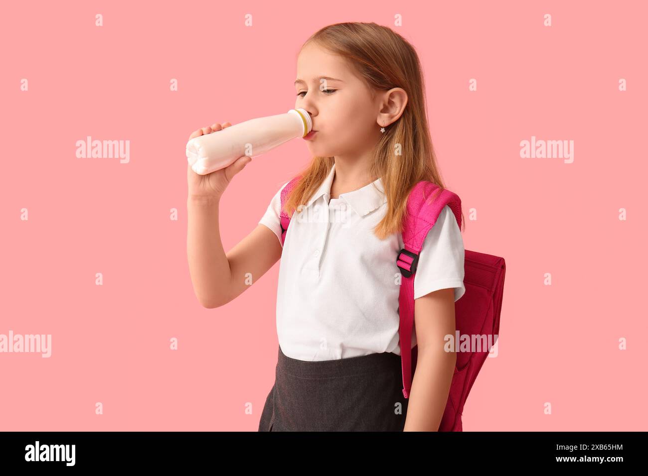Cute little schoolgirl with backpack drinking yogurt on pink background ...