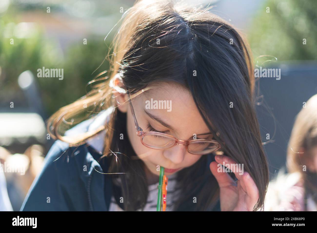 Little sad girl in glasses sits at a table outdoors drinking through a ...